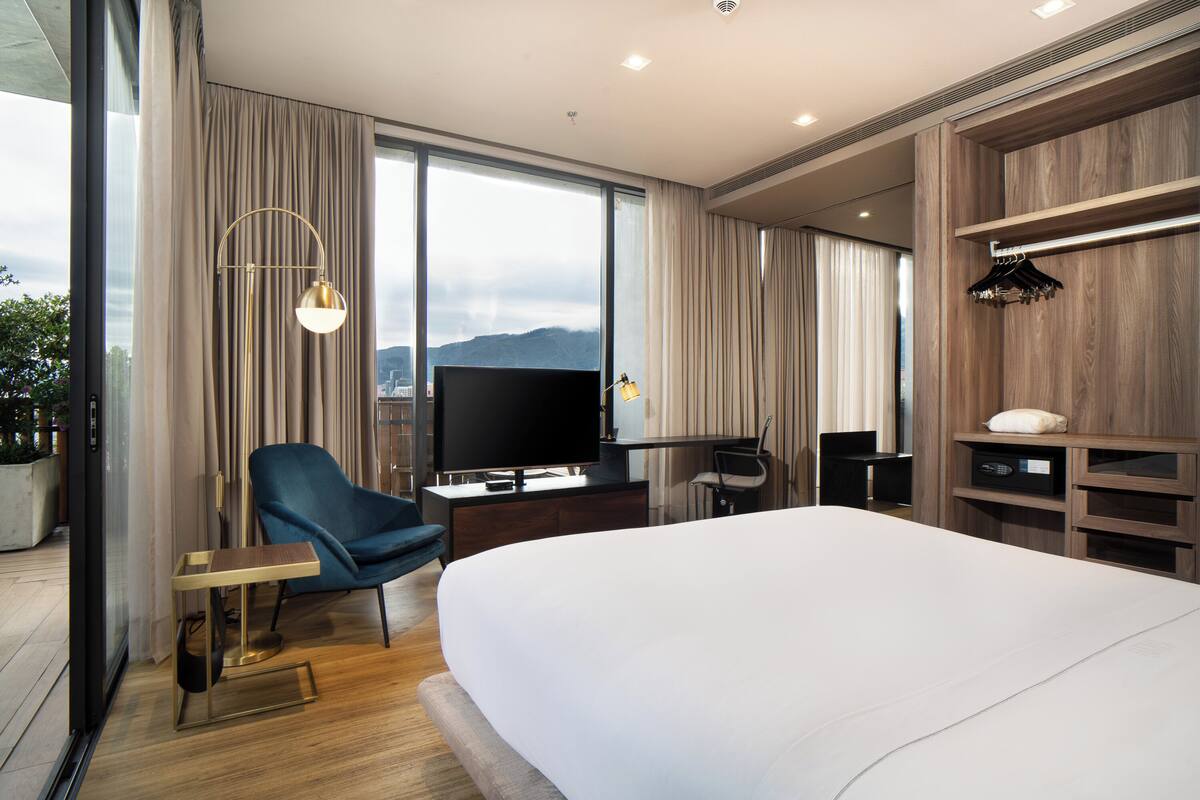 Quarto de hotel moderno com uma cama grande, uma escrivaninha com televisão de tela plana, uma poltrona azul e vista da cidade através de grandes janelas.