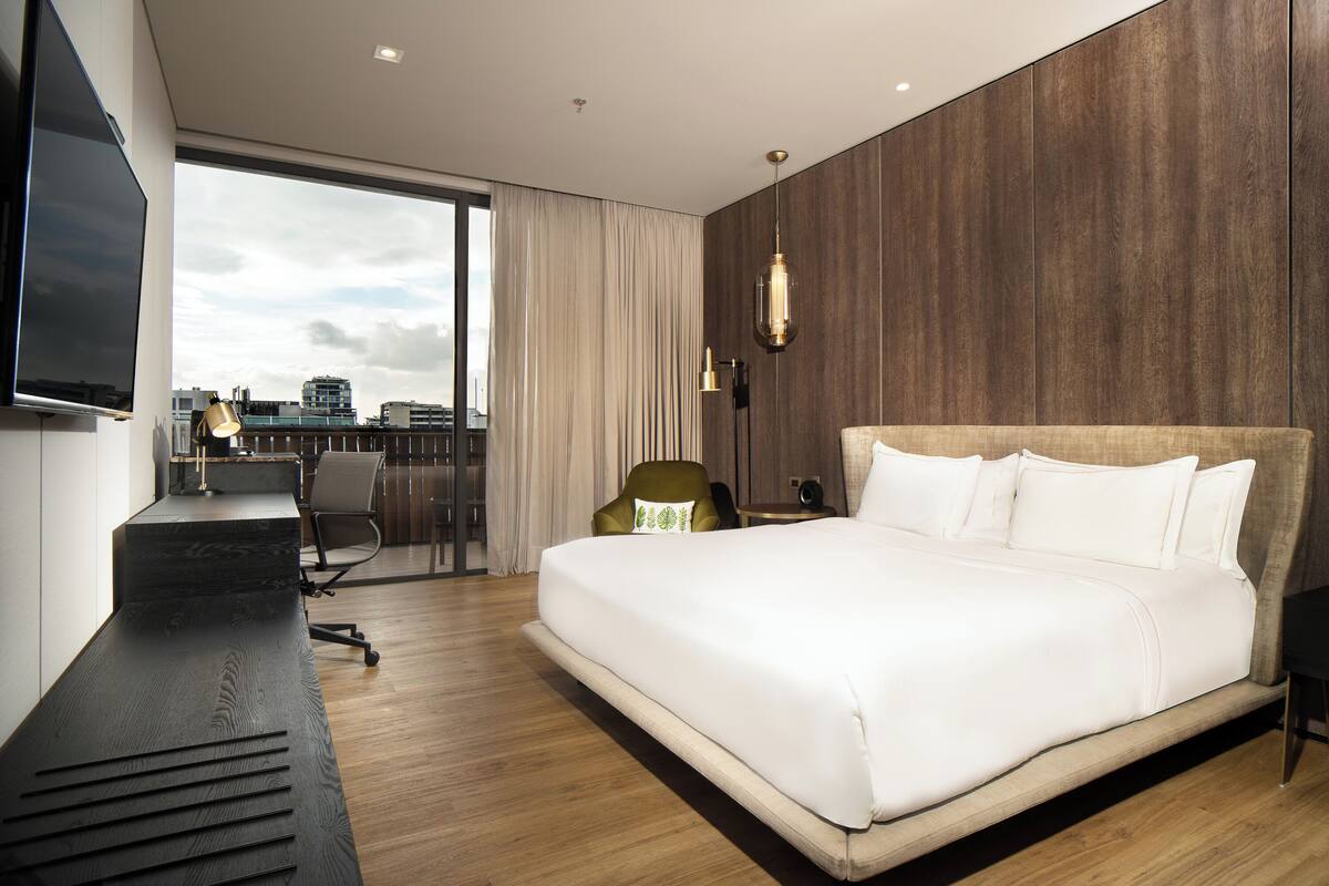 Quarto de hotel moderno com uma cama grande, uma escrivaninha, uma cadeira, uma televisão e vista para a cidade.