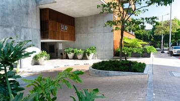 Um edifício moderno com fachada de concreto e painel de madeira no nível superior. Há plantas em vasos e uma área ajardinada com vegetação.