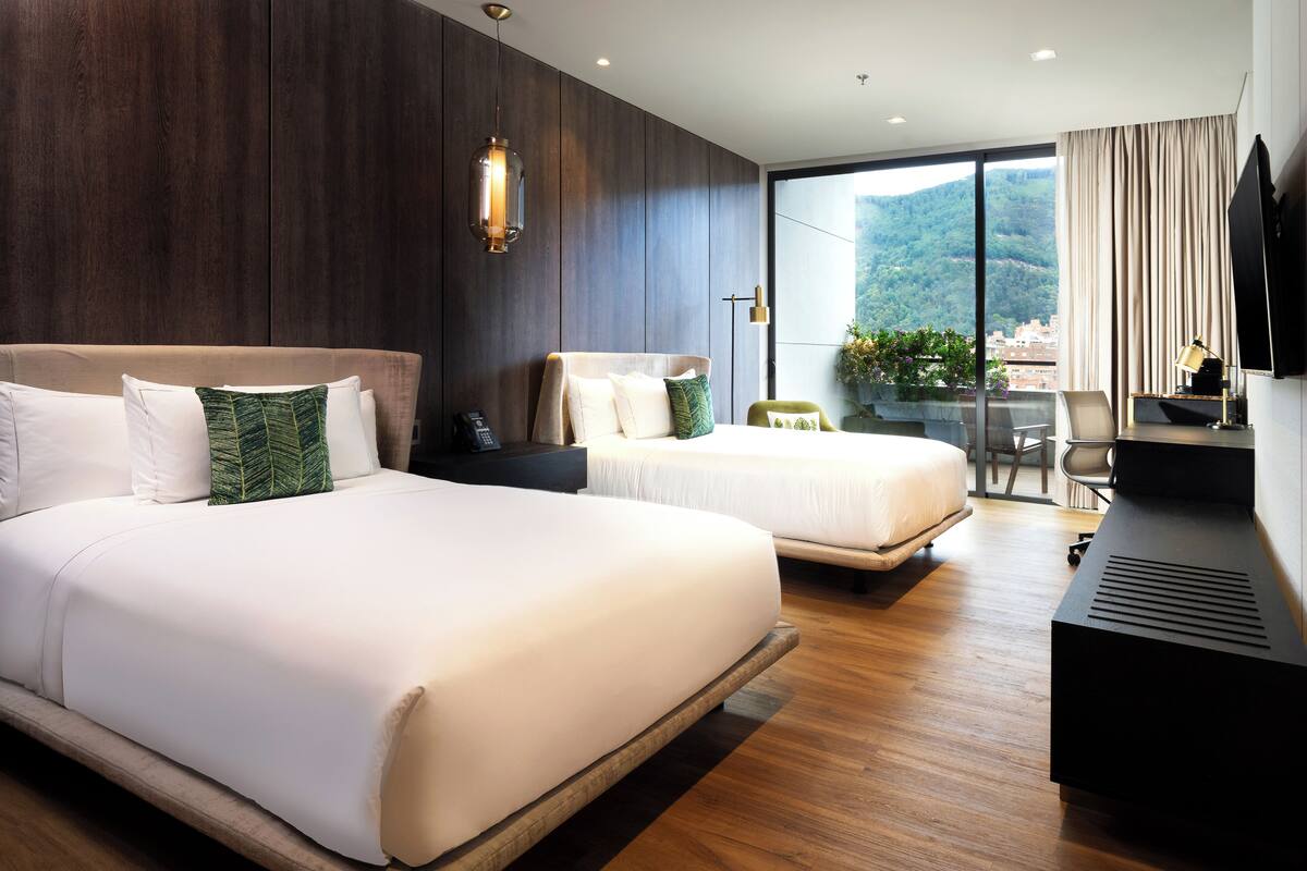 Quarto de hotel moderno com duas camas, uma mesa de trabalho e uma janela grande com vista para a cidade.