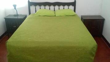 Uma cama com colcha verde, duas almofadas, cabeceira de madeira e mesa de cabeceira com abajur.
