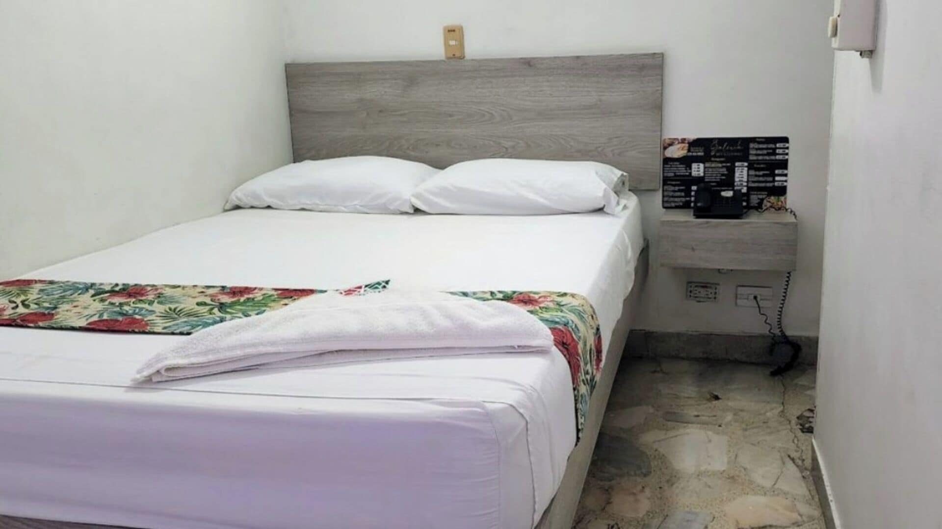 Uma cama pequena e bem arrumada, com lençóis brancos e manta estampada com flores, cabeceira de madeira, mesa de cabeceira com um objeto preto e chão com padrão de pedra.