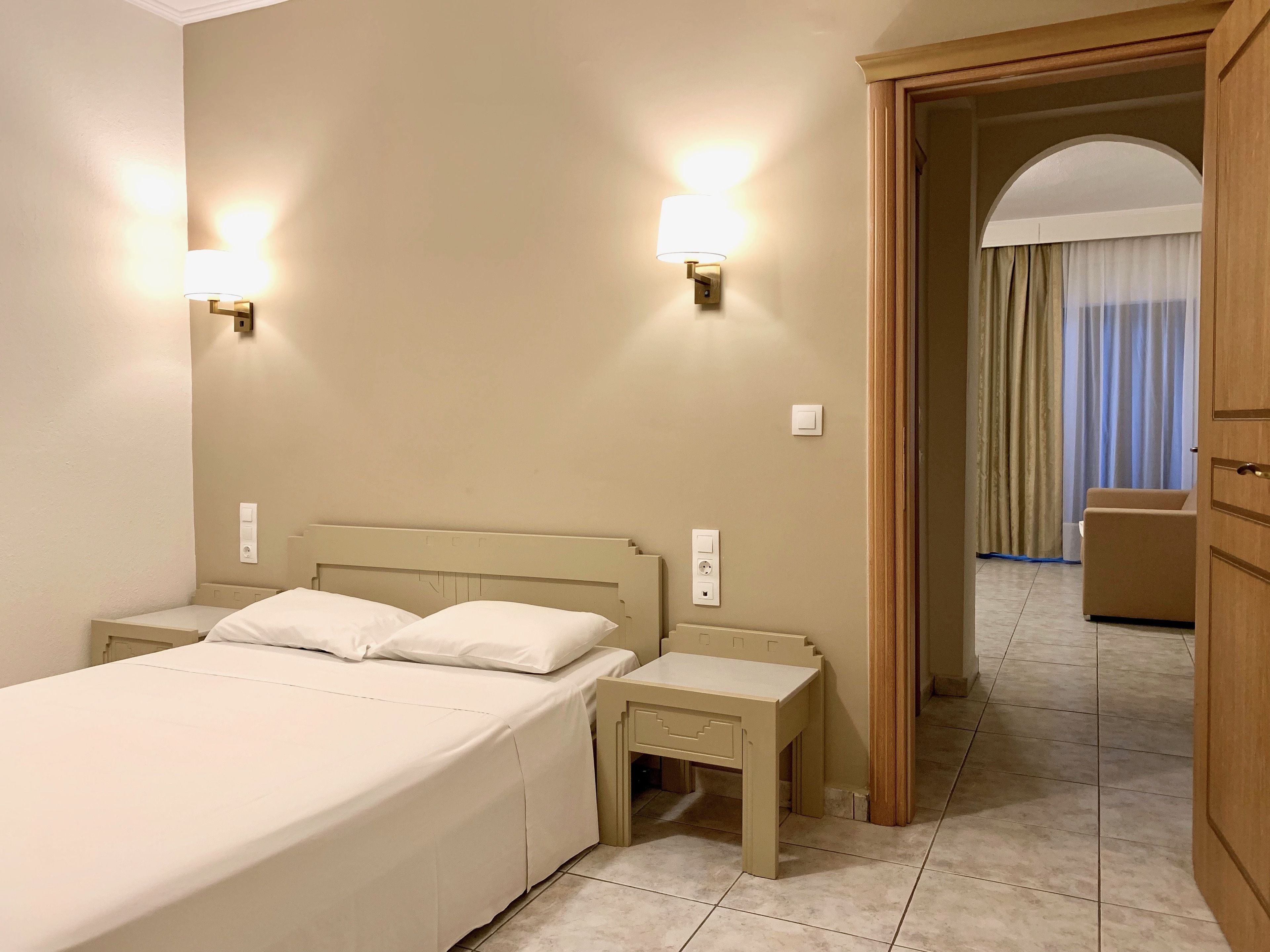 Quarto de hotel com cama, mesas de cabeceira, luminárias fixadas na parede, guarda-roupa de madeira e vista para uma varanda com cortinas.