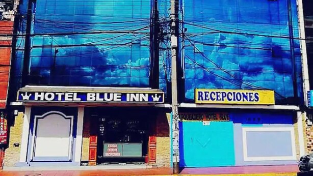 Hotel com fachada de vidro azul refletindo o céu, área de recepção e placa com a inscrição 'Hotel Blue Inn'.