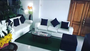 Uma sala de estar com um sofá branco, almofadas escuras, uma mesa de centro e um tapete verde.