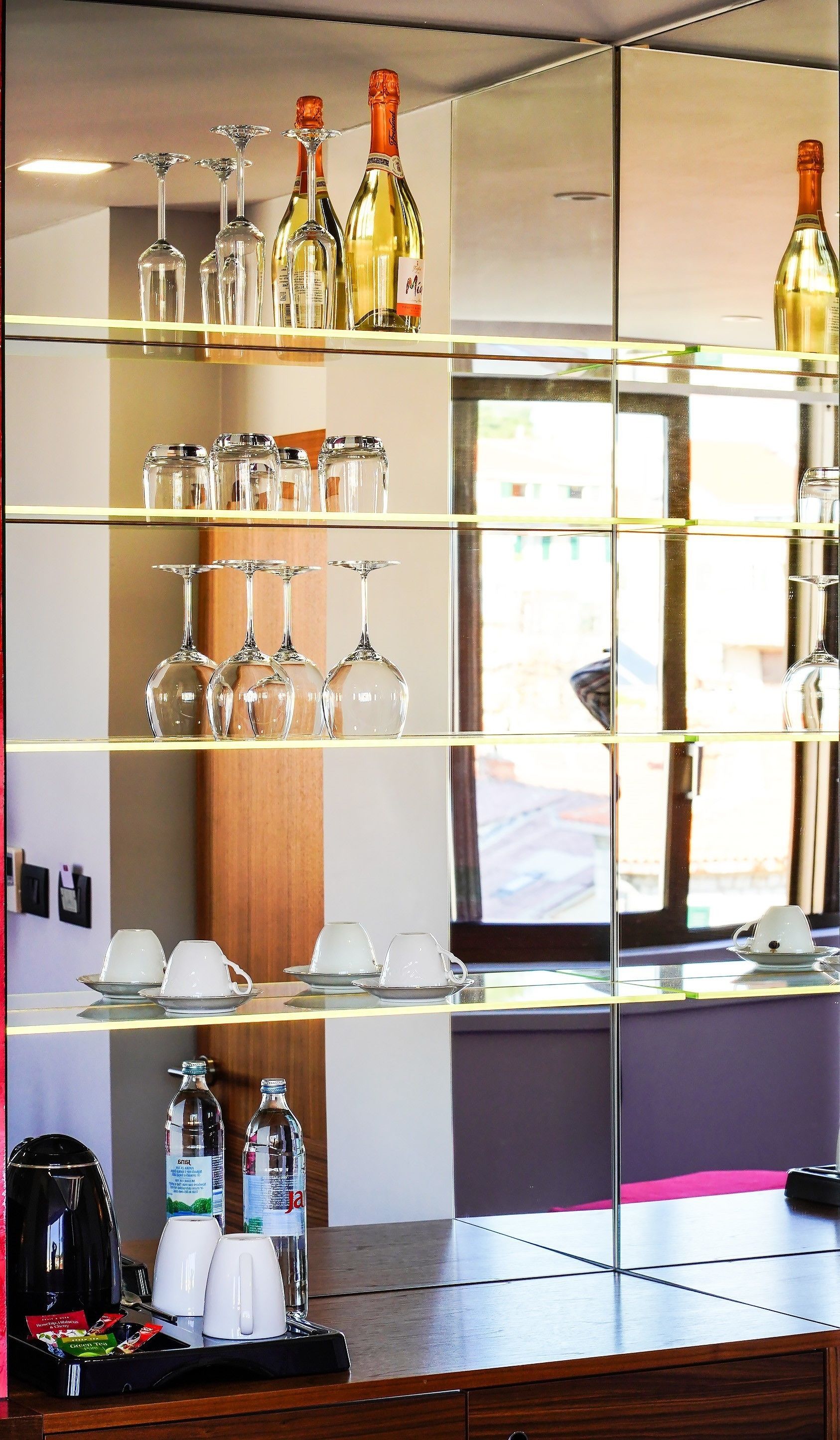 Quarto de hotel com minibar, espaço para café e vitrine de vidro com garrafas de vinho e copos.