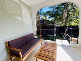 Varanda com um banco de madeira e um sofá escuro, com vista para um jardim com árvores e cerca de madeira.