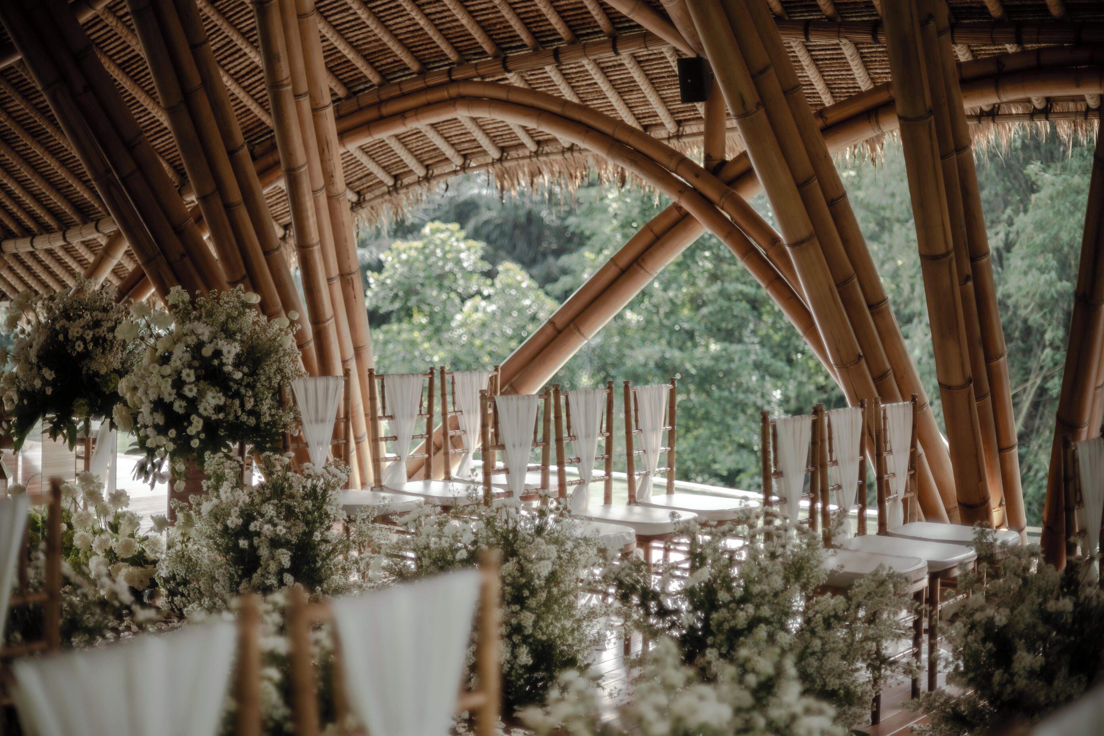 Espaço interno com estruturas de bambu, cadeiras brancas e decoração floral.