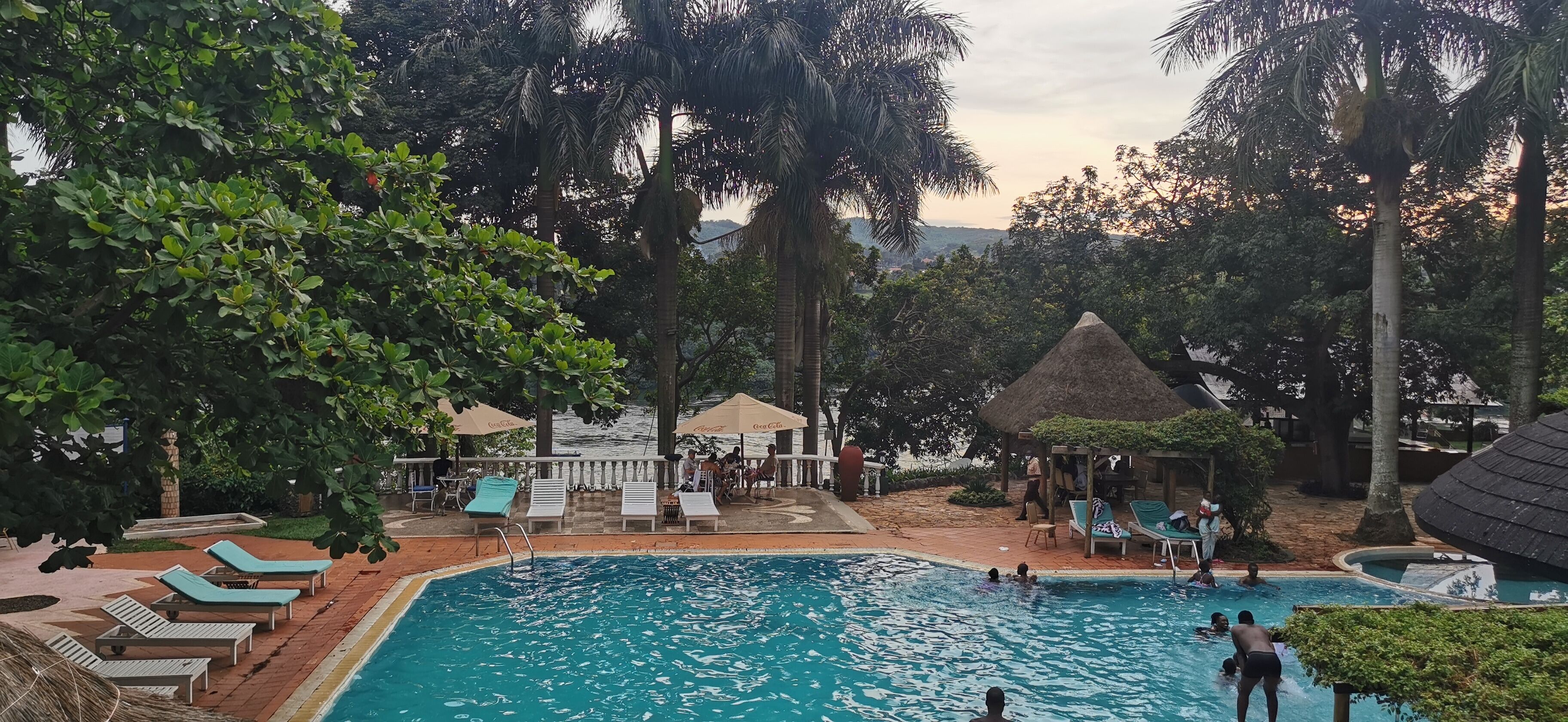 Uma área de piscina com espreguiçadeiras, uma estrutura com telhado de palha e vegetação ao redor.