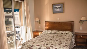 Quarto com cabeceira de madeira, colcha florida, duas mesas de cabeceira com luminárias e vista para uma varanda com grade de treliça.
