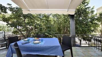 Uma área de refeições ao ar livre coberta, com uma mesa posta para dois, rodeada de cadeiras e vista para árvores e edifícios ao fundo.