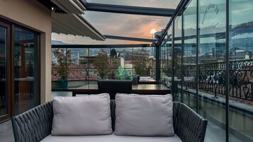 Uma área moderna de estar ao ar livre com estrutura de vidro, proporcionando vista para a cidade e um pôr do sol.