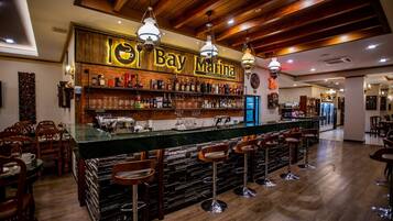 Uma área de bar com teto de madeira, balcão com banquetas e prateleiras repletas de garrafas.