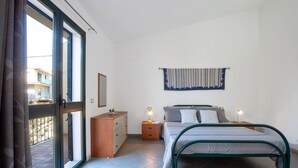 Quarto com cama, mesa de cabeceira, cômoda, janela com cortinas e vista para o exterior.