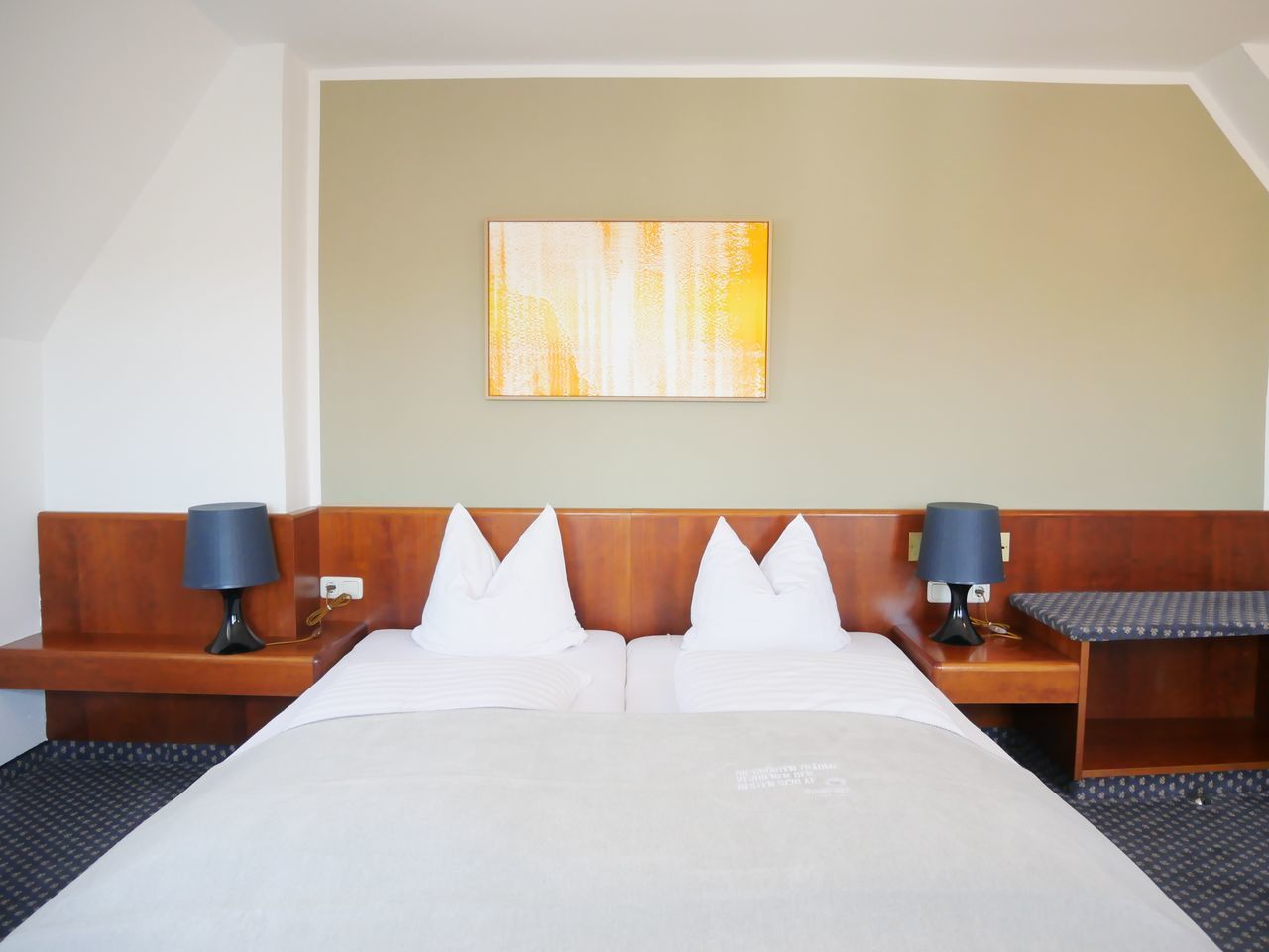 Quarto de hotel com uma cama grande, duas mesas de cabeceira com luminárias, uma cabeceira e uma obra de arte na parede.