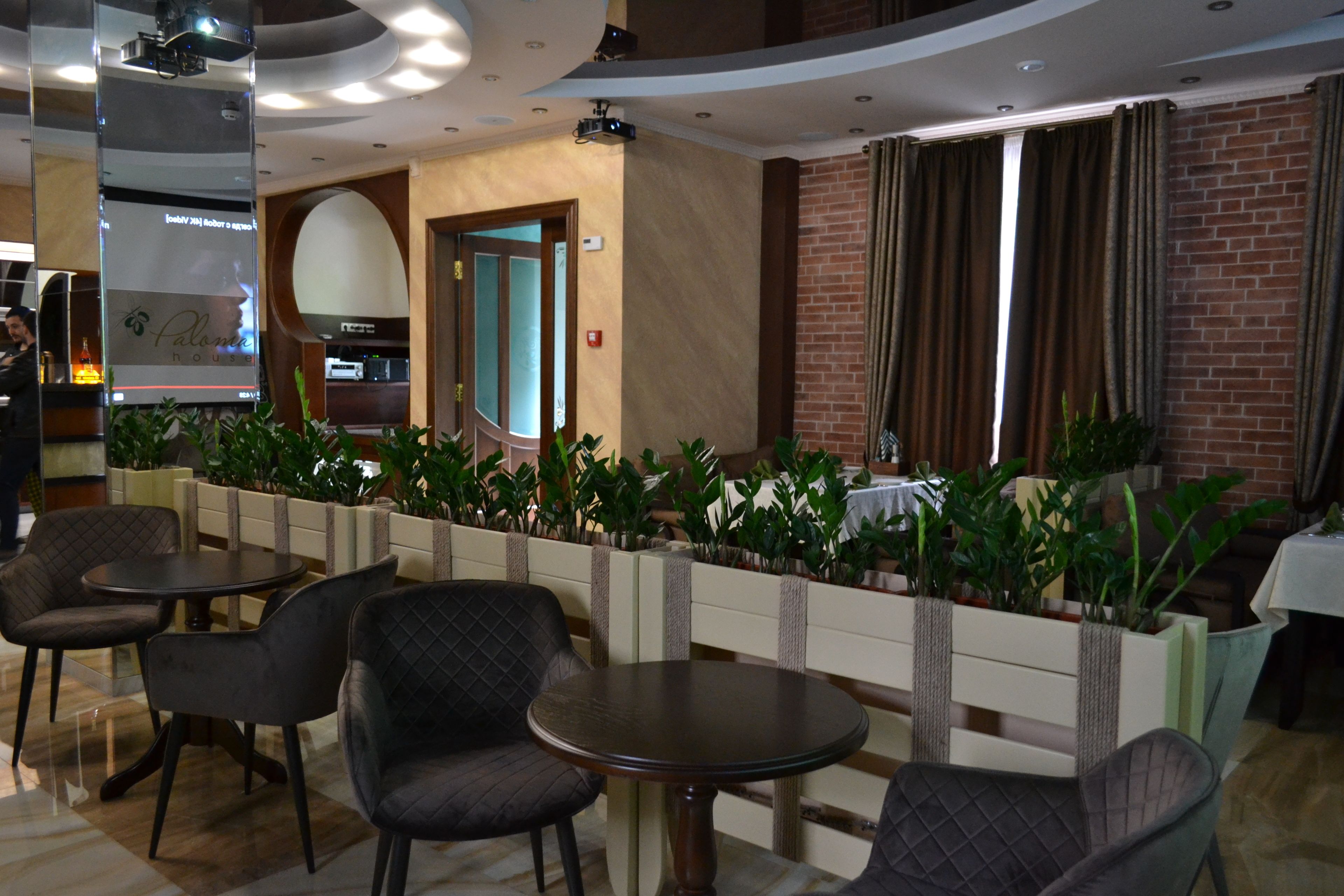 Uma sala de jantar moderna, com mesas redondas e cadeiras estofadas, parede de tijolos e uma barreira vegetal.