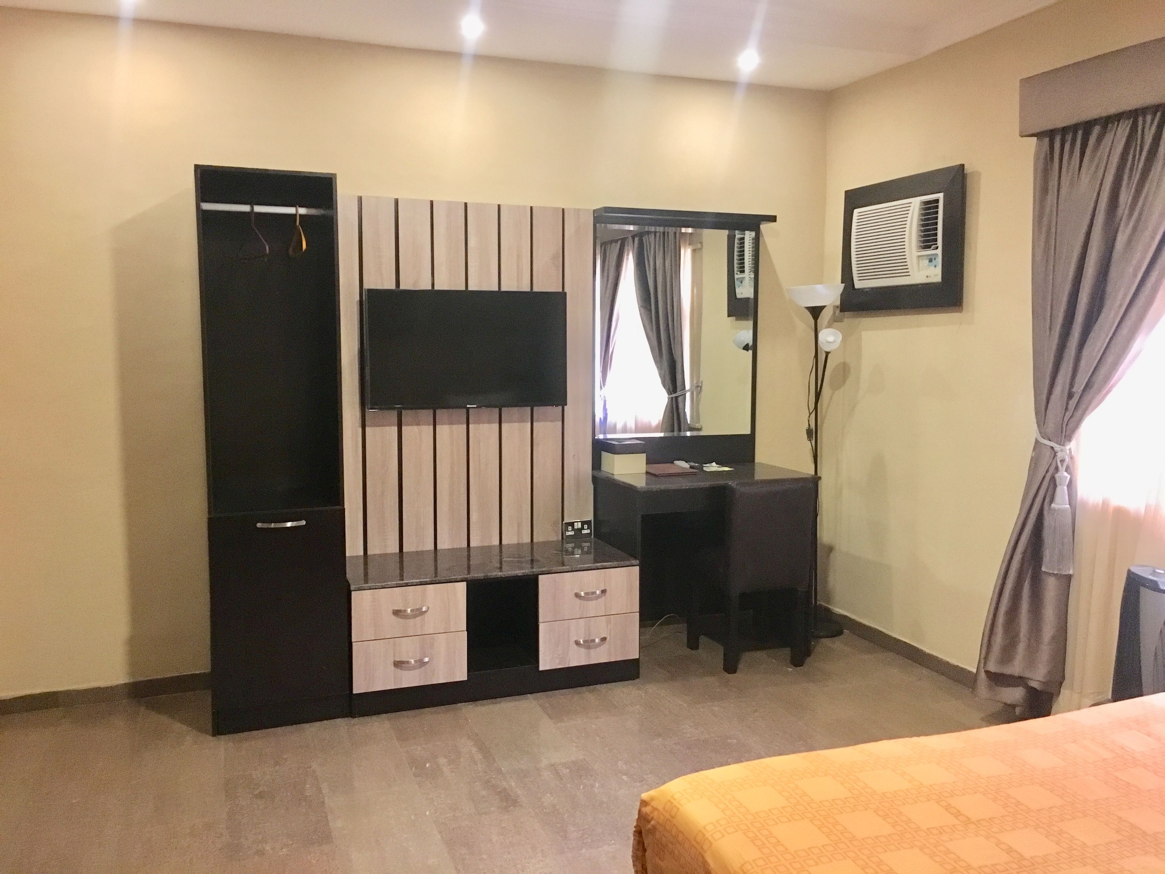 Quarto de hotel com cama, televisão na parede, secretária com gavetas, espelho e janela com cortinas.