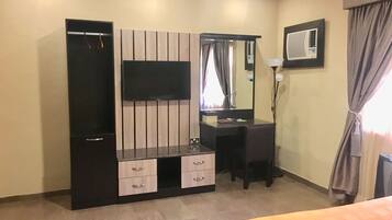 Quarto de hotel com cama, televisão na parede, secretária com gavetas, espelho e janela com cortinas.