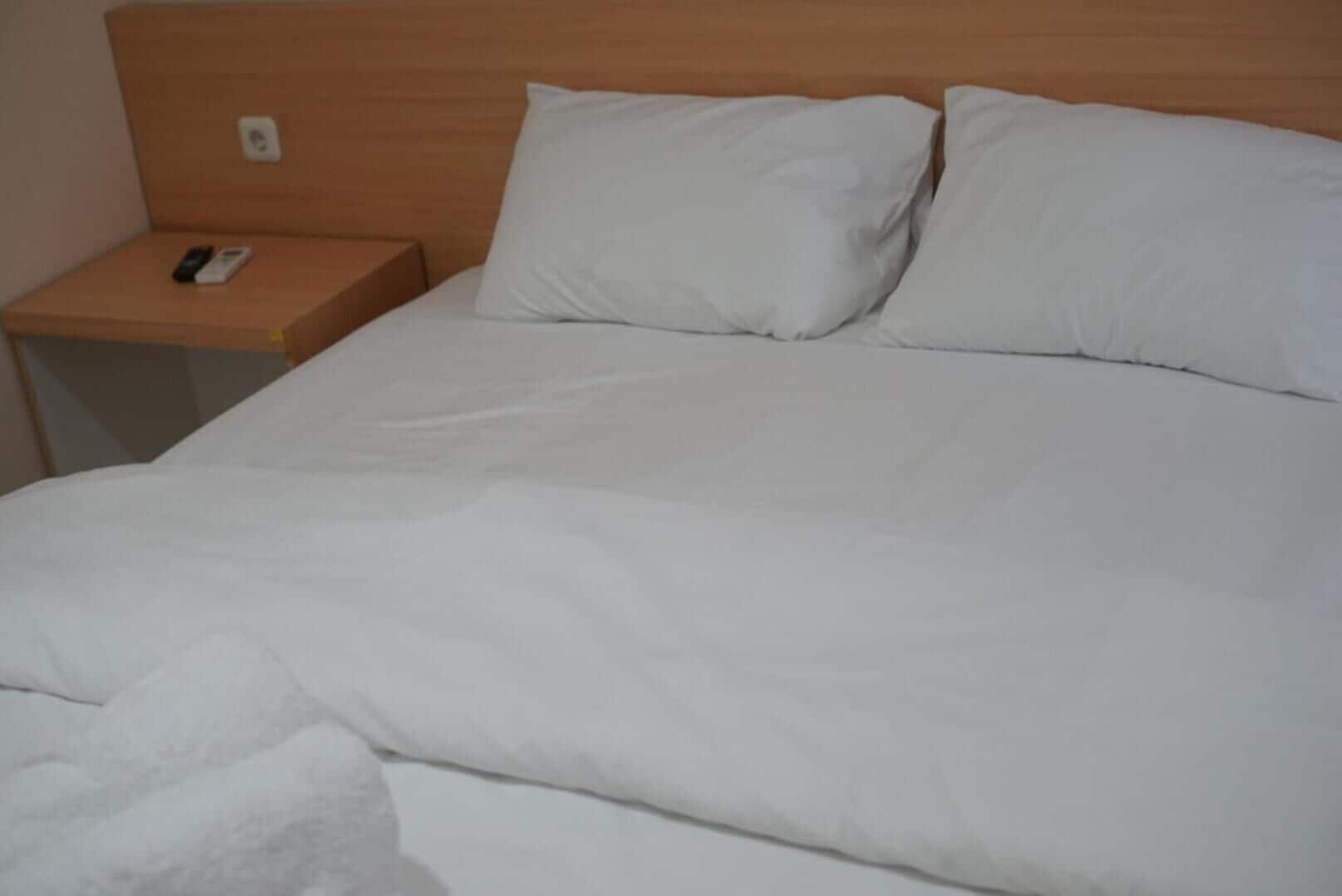 Uma cama com roupa de cama branca, cabeceira de madeira e mesa de cabeceira com controle remoto.