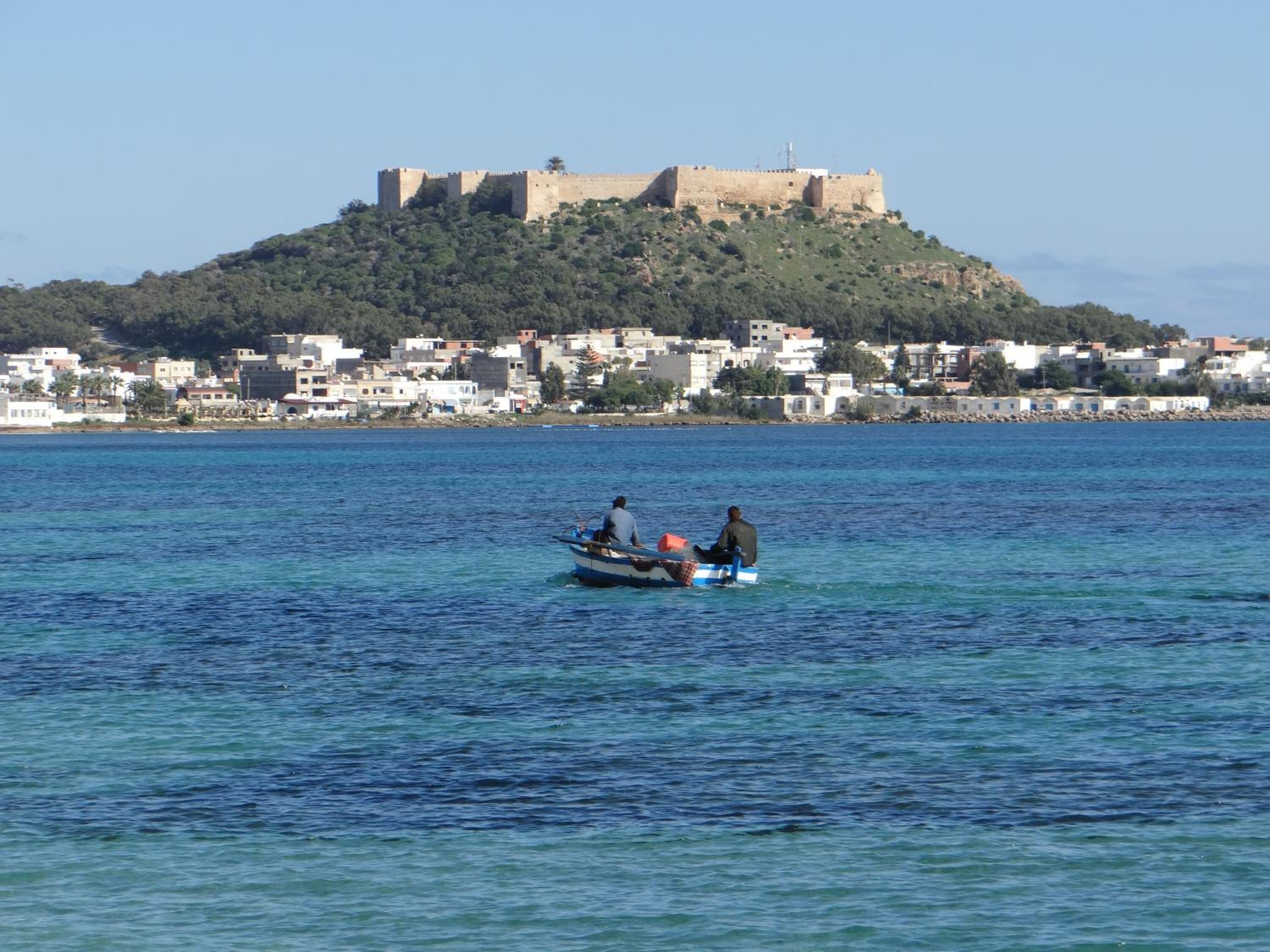 Uma vila costeira com uma fortaleza histórica numa colina, mar calmo e pequenos barcos com pessoas.