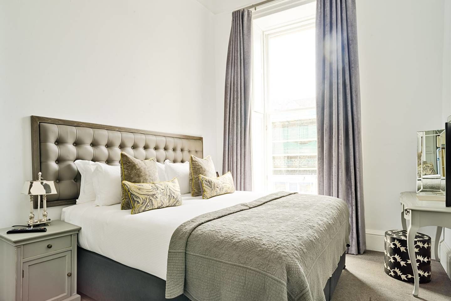Quarto com cabeceira estofada, cama com roupa de cama branca e almofadas estampadas, mesa de cabeceira com abajur e uma janela grande com cortinas.
