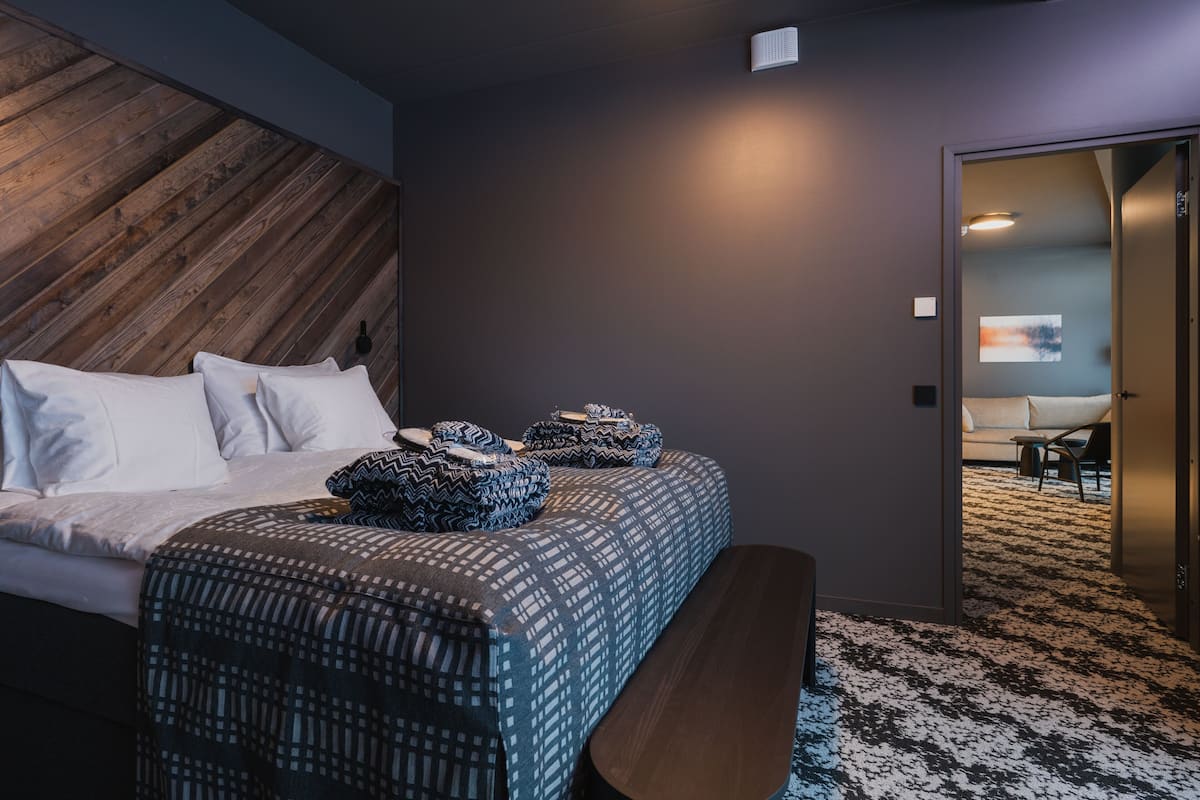 Quarto de hotel com uma cama, um banquinho, cabeceira de madeira, colcha estampada e piso coberto de carpete.