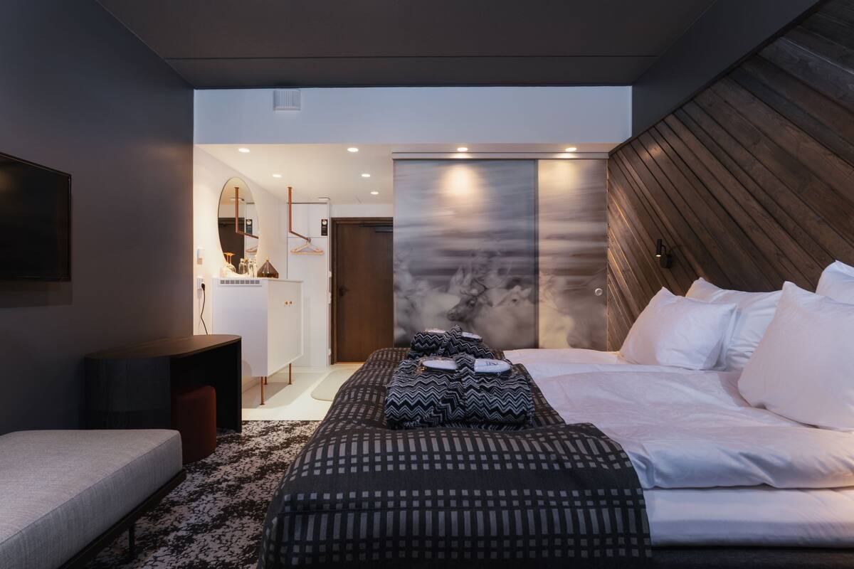 Quarto de hotel moderno com uma cama grande, uma mesinha de cabeceira, um pequeno espaço de estar e um guarda-roupa.