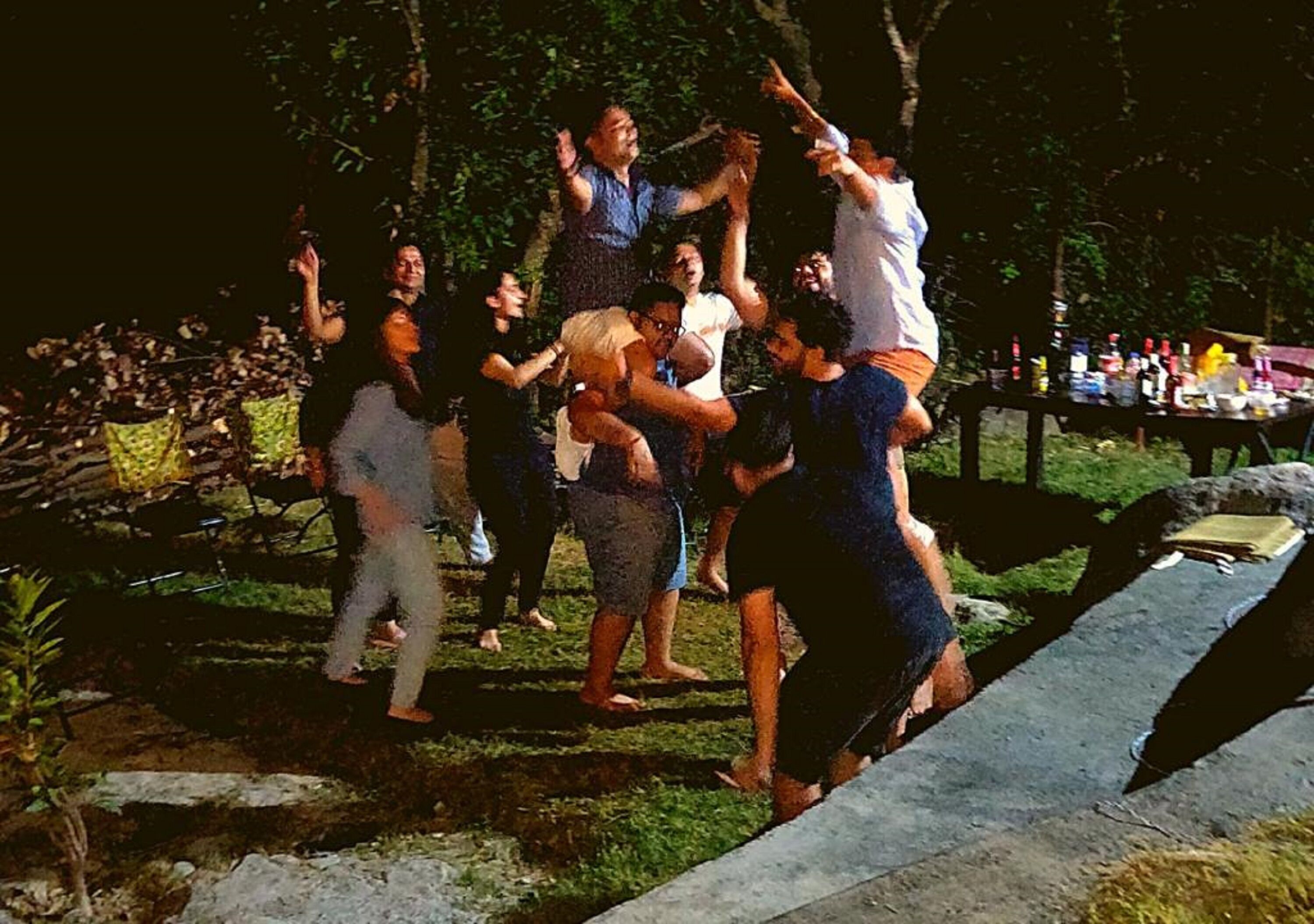 Um grupo de pessoas dançando ao ar livre em um ambiente natural, com uma mesa de bebidas e petiscos.
