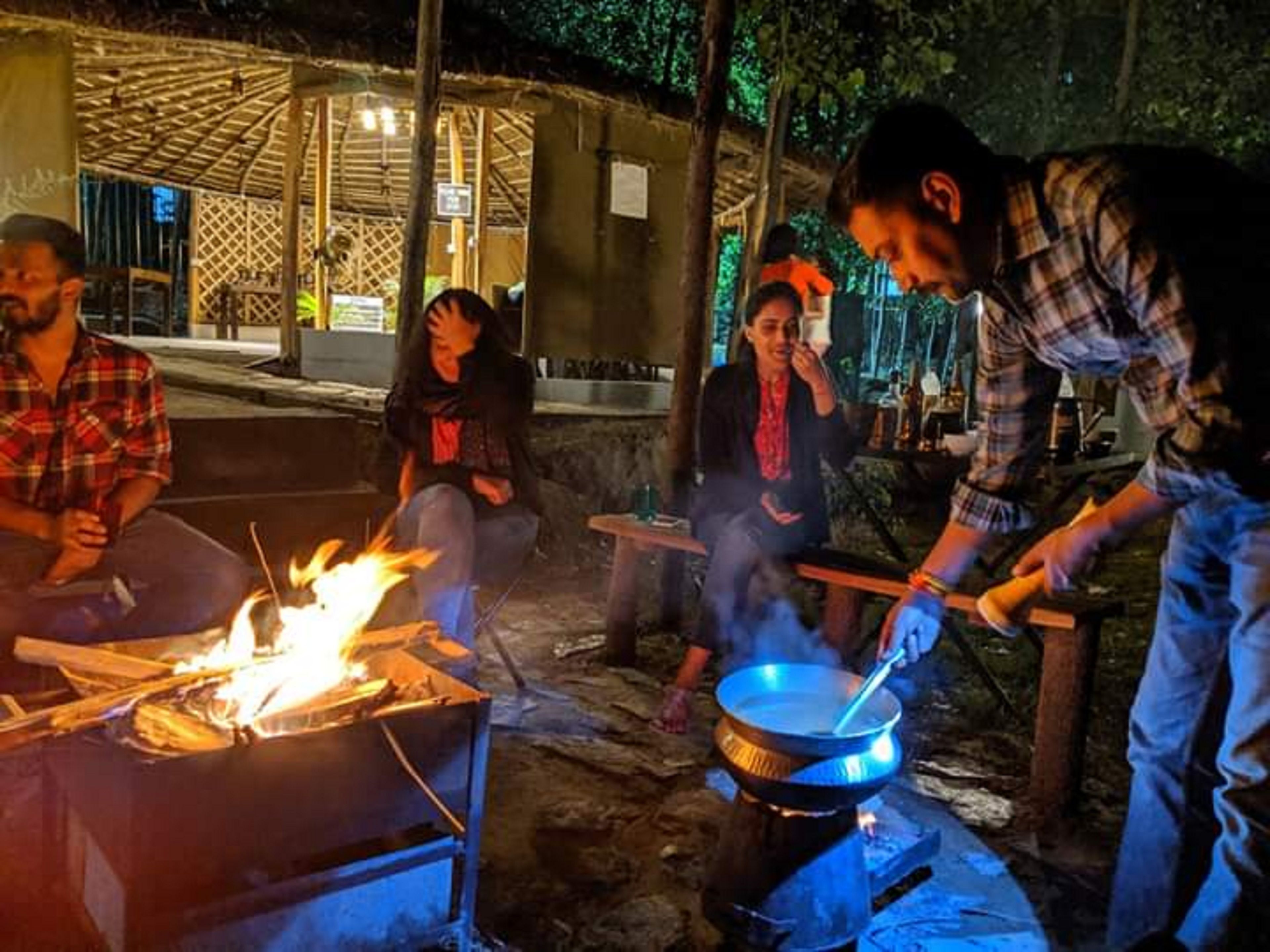 Um grupo de pessoas reunido em volta de uma fogueira, com uma panela esquentando em um fogão portátil.