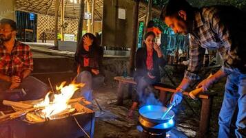 Um grupo de pessoas reunido em volta de uma fogueira, com uma panela esquentando em um fogão portátil.