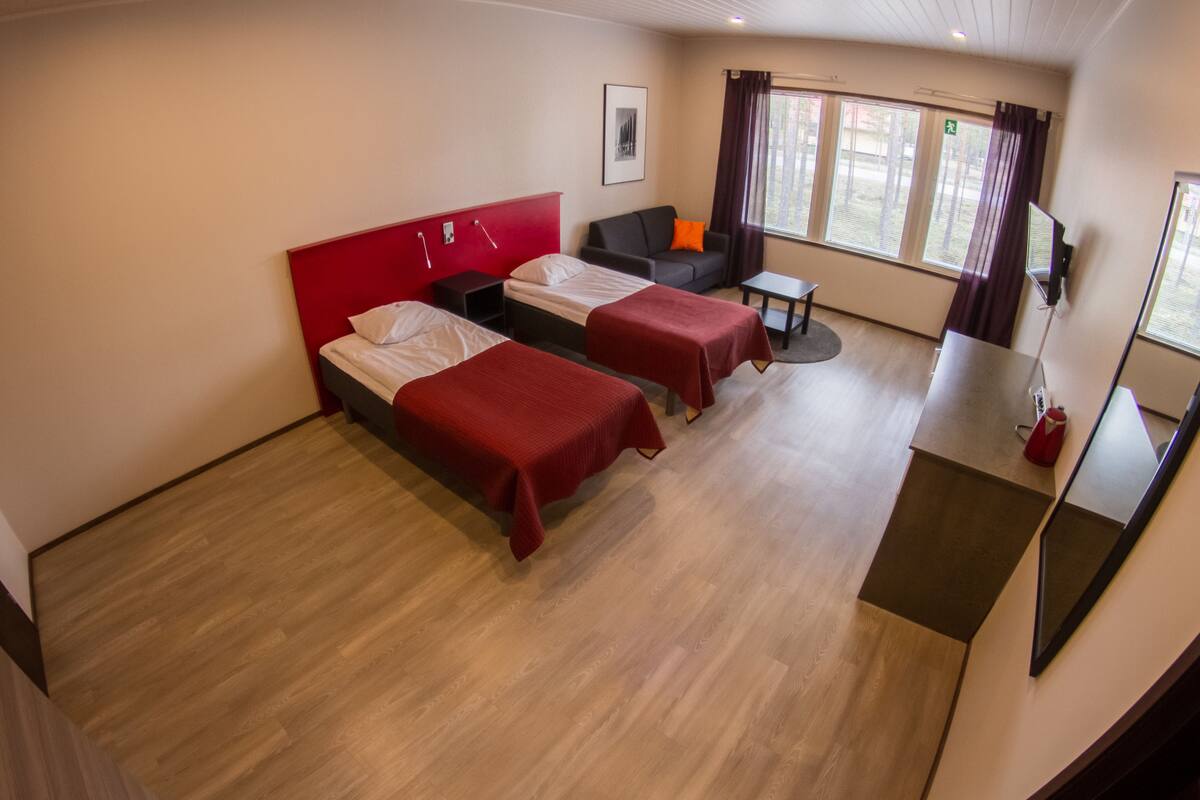 Quarto de hotel com duas camas de solteiro, um sofá, uma mesinha e uma televisão.