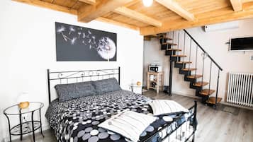 Quarto com cama, edredom preto e branco com bolinhas, cabeceira de metal preta, mesa redonda pequena com abajur, arte na parede em preto e branco, escada e radiador branco.