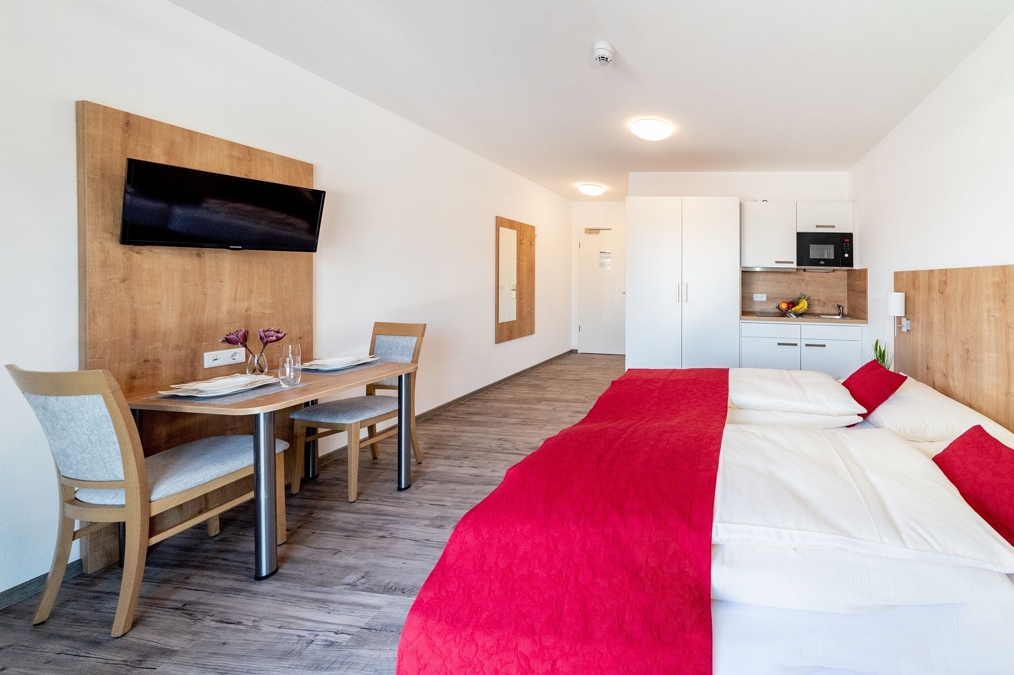 Quarto de hotel com uma cama, uma escrivaninha com cadeiras, uma televisão na parede, um micro-ondas e uma kitchenette pequena.
