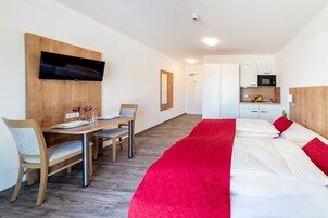 Quarto de hotel com uma cama, uma escrivaninha com cadeiras, uma televisão na parede, um micro-ondas e uma kitchenette pequena.