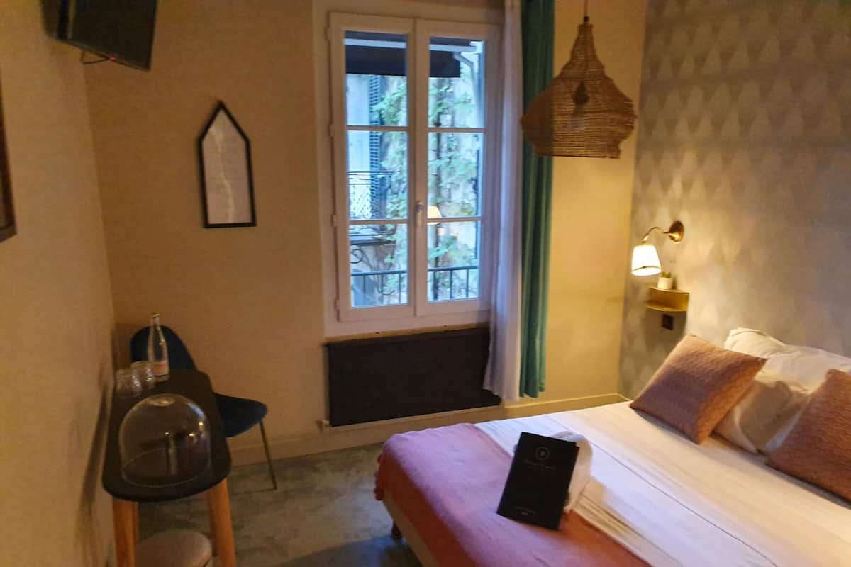 Quarto de hotel com cama, escrivaninha, cadeira, janela e luminária na parede.