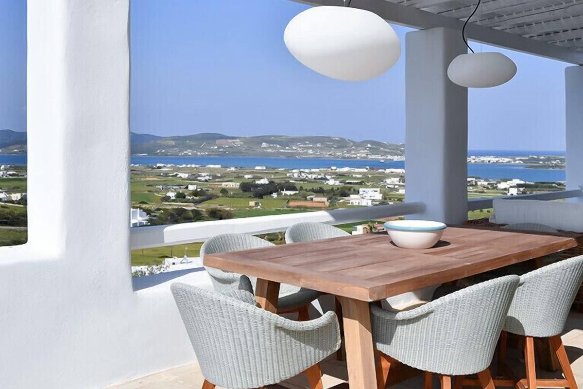 Mesa de madeira com cadeiras e uma tigela, com vista para uma paisagem costeira com casas e o mar.