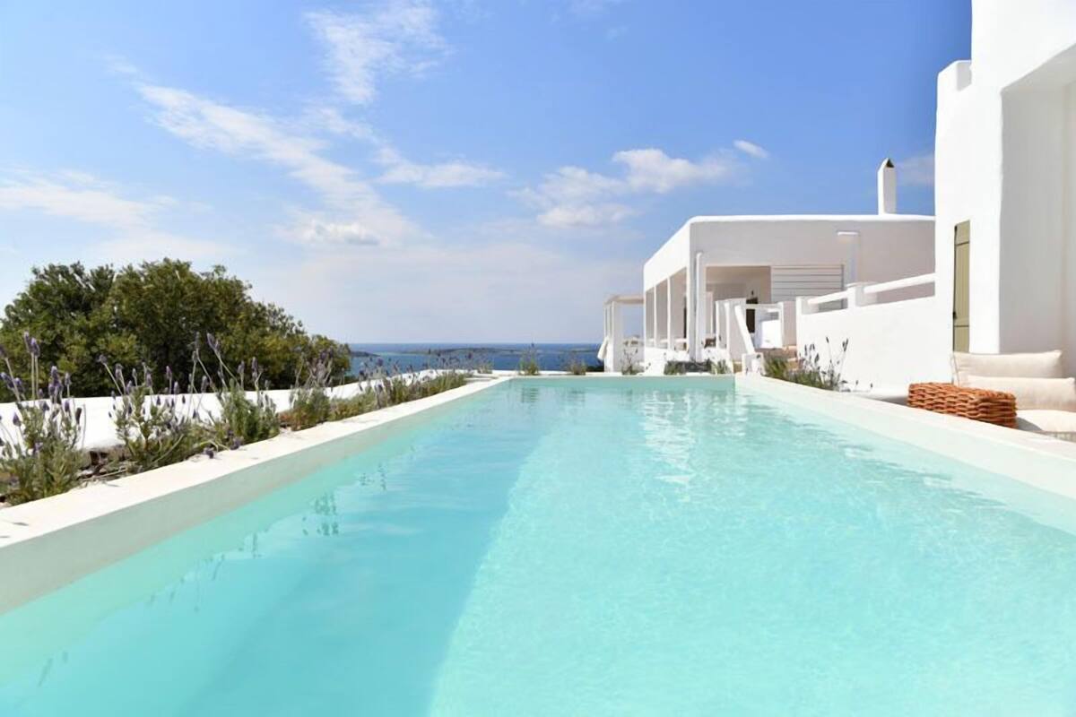 Uma piscina comprida e retangular com água azul cristalina, rodeada por edifícios brancos e vista para o mar.