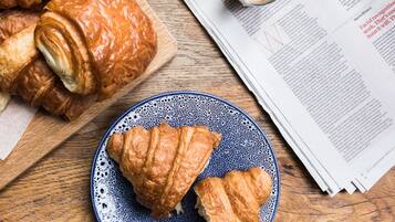 Uma mesa de madeira com croissants, um prato com um croissant, uma chávena de café e um jornal.