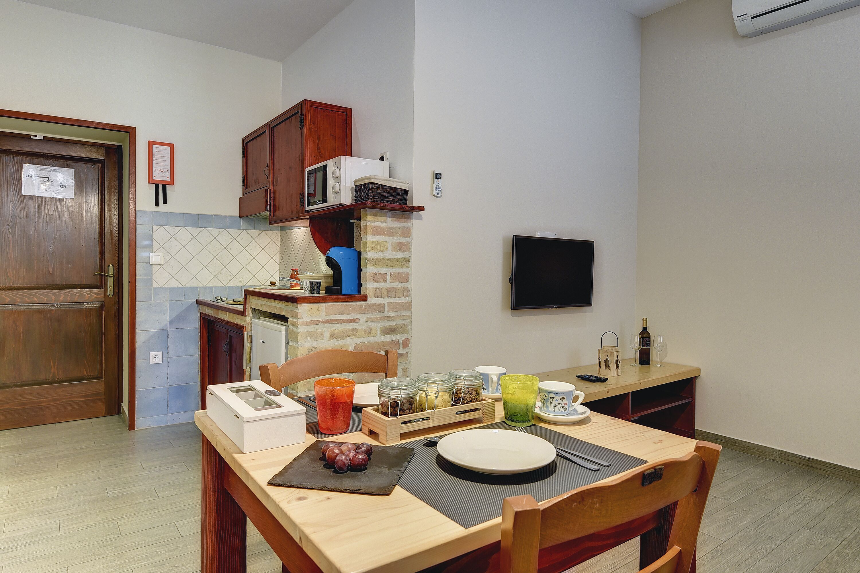 Uma cozinha compacta com micro-ondas, um frigorífico pequeno e um armário de madeira. Há uma mesa de jantar com cadeiras, uma televisão fixada na parede e uma porta com um aviso.