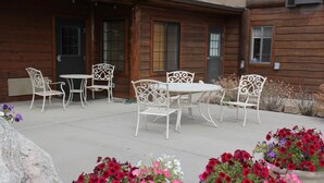 Uma cabana de madeira com área de varanda, com cadeiras e mesas de metal branco, rodeada por canteiros de flores vermelhas e cor-de-rosa.
