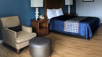 Quarto de hotel com cama, cadeira, mesa de cabeceira, telefone e um pequeno banquinho.