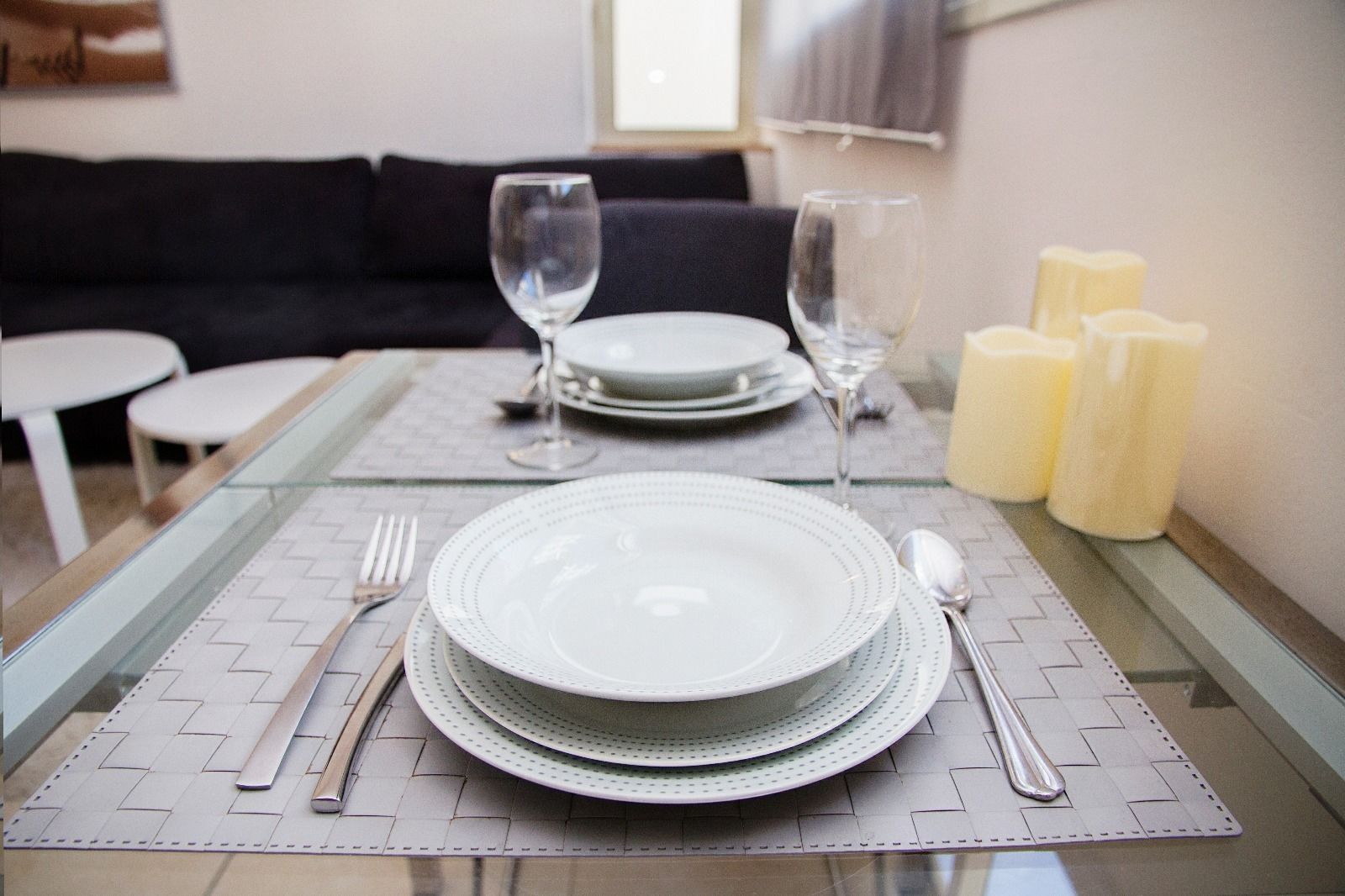 Uma mesa de jantar posta com pratos, talheres e velas, com um sofá e cadeiras ao fundo.