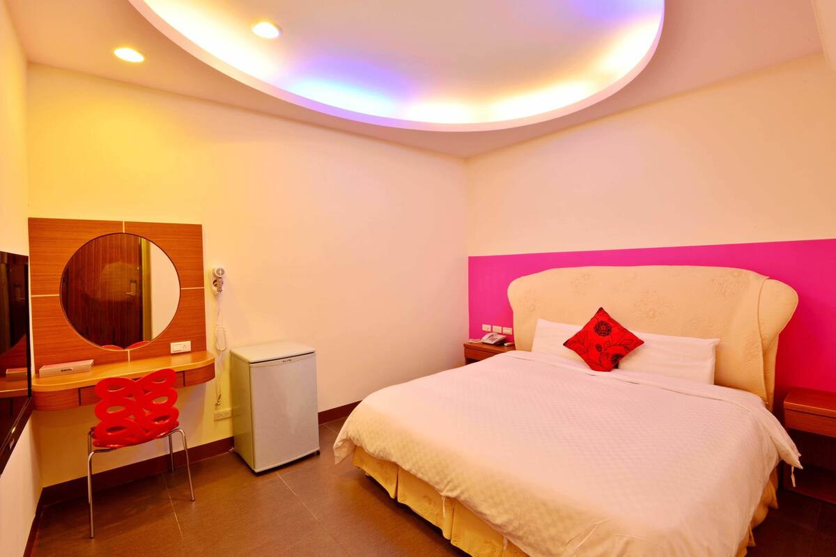 Quarto de hotel moderno com cama, escrivaninha, cadeira, um pequeno frigorífico e espelho.