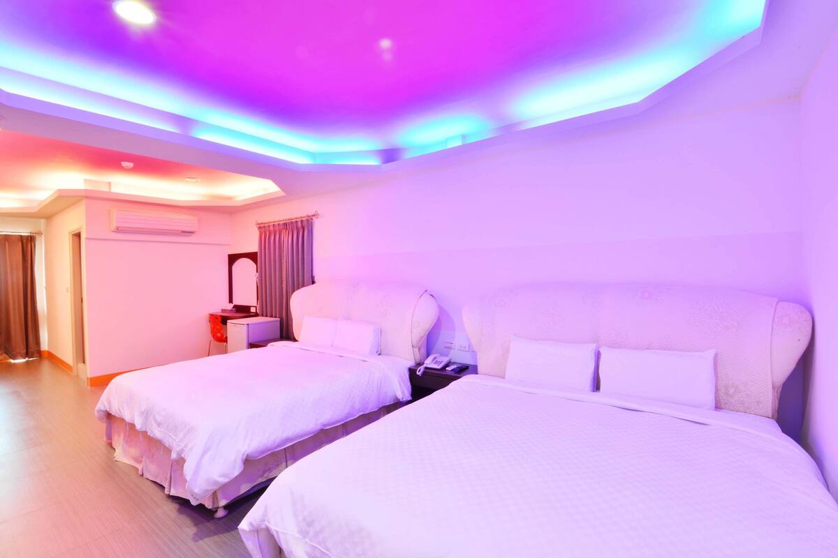 Quarto de hotel com duas camas, uma mesa de cabeceira, um espelho e iluminação colorida no teto.