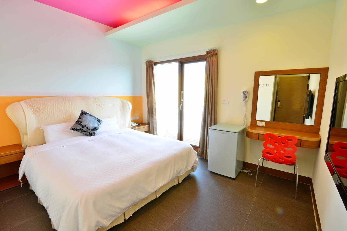 Quarto de hotel com uma cama, escrivaninha, cadeira, espelho e porta.