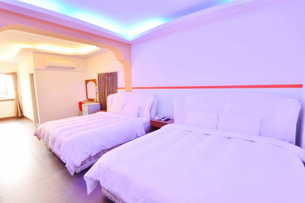 Quarto de hotel com duas camas, uma mesinha de cabeceira, um espelho e uma janela com cortinas.