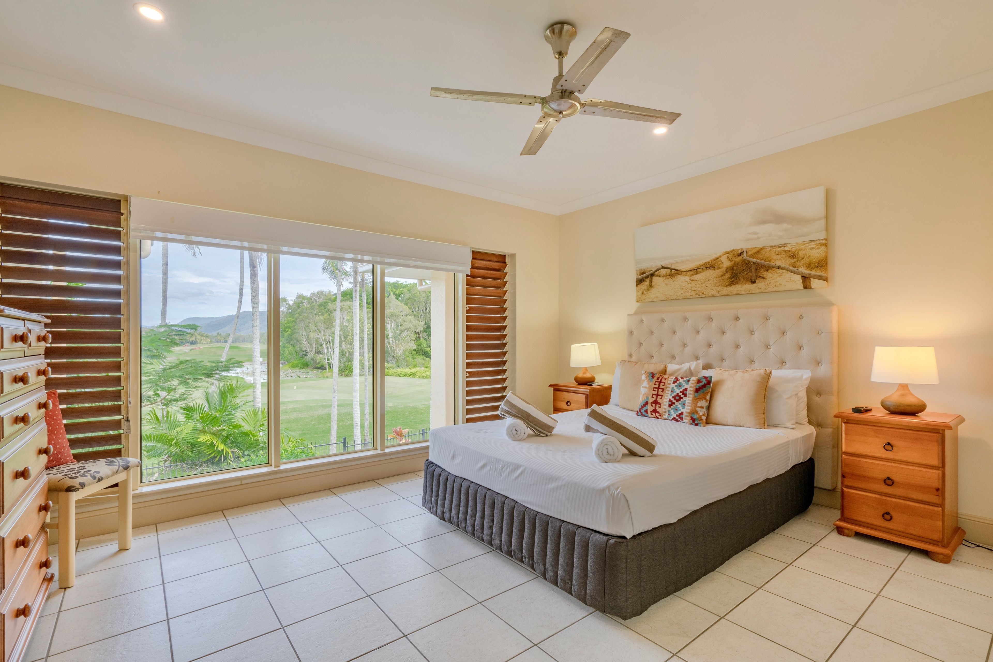 Quarto com uma cama grande, um ventilador de teto, uma mesa de cabeceira com abajur e vista para uma paisagem verde pela janela.