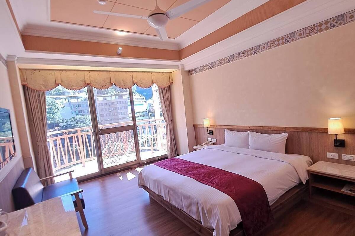 Quarto de hotel com uma cama grande, um ventilador de teto, mesa de cabeceira com abajur e vista para a varanda.