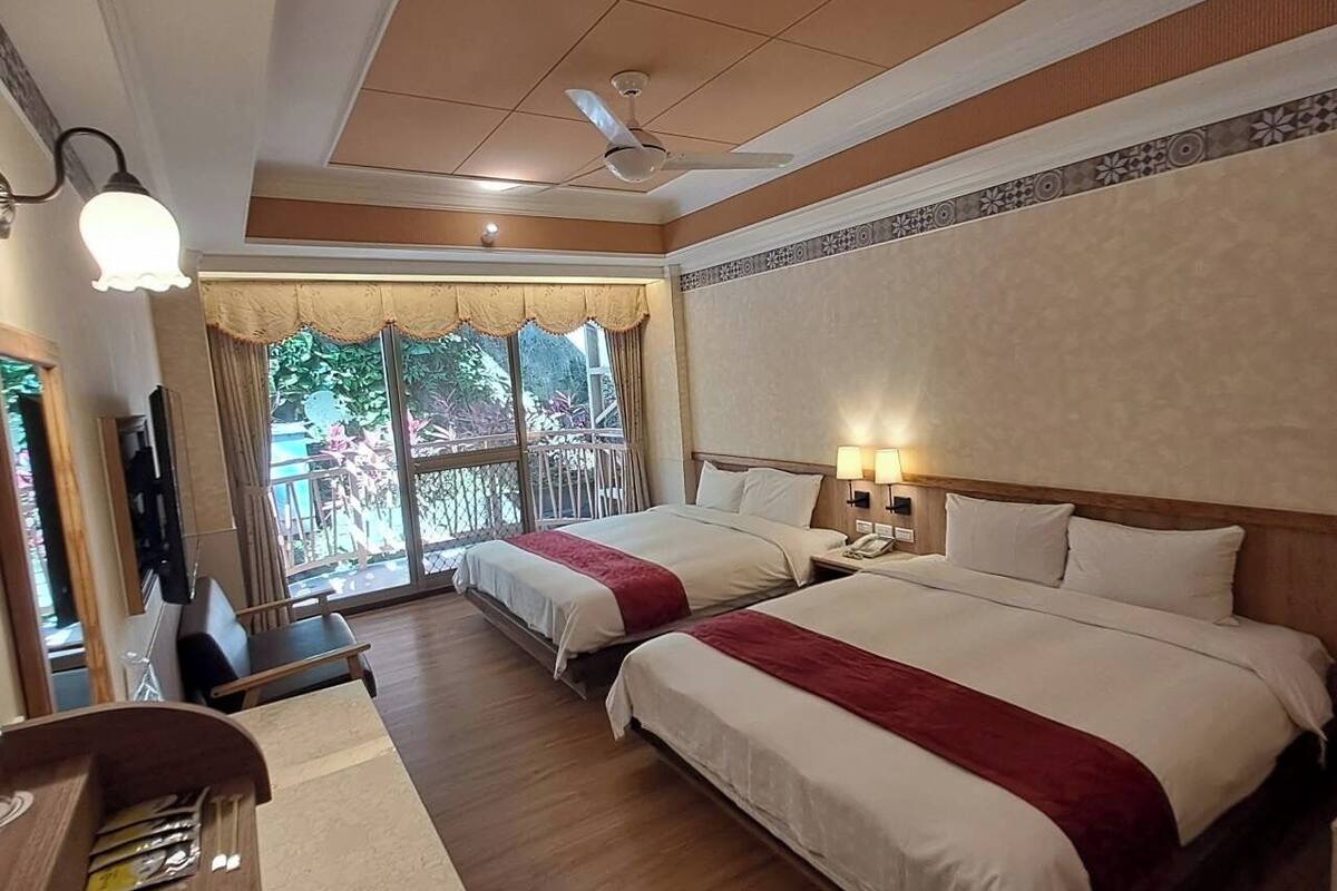 Quarto de hotel com duas camas, uma escrivaninha, uma cadeira, uma televisão e varanda com vista para o verde.