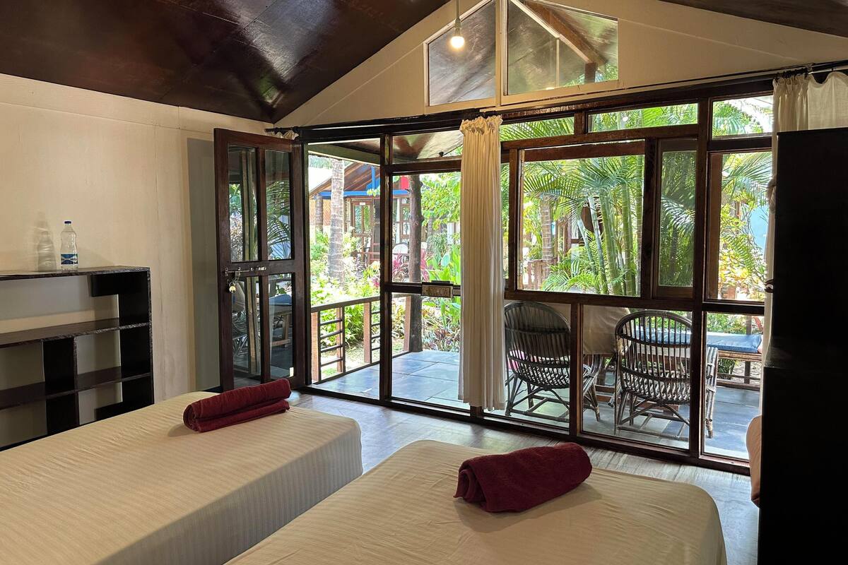 Quarto com duas camas, uma escrivaninha e amplas janelas com vista para o verde lá fora.